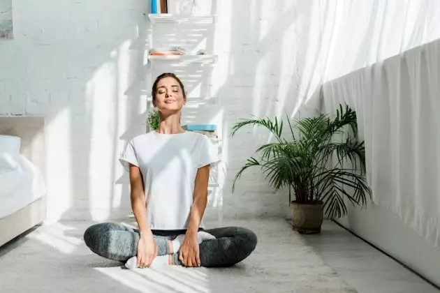 Tetap sehat dengan anggaran terbatas: gadis cantik yang bahagia dengan mata tertutup berlatih yoga dalam posisi lotus di kamar tidur di pagi hari