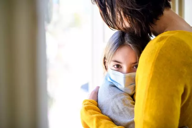 Informasi yang salah tentang vaksin: Gadis sekolah kecil dengan topeng wajah dan ibu yang tidak dapat dikenali di dalam rumah di rumah, berpelukan.